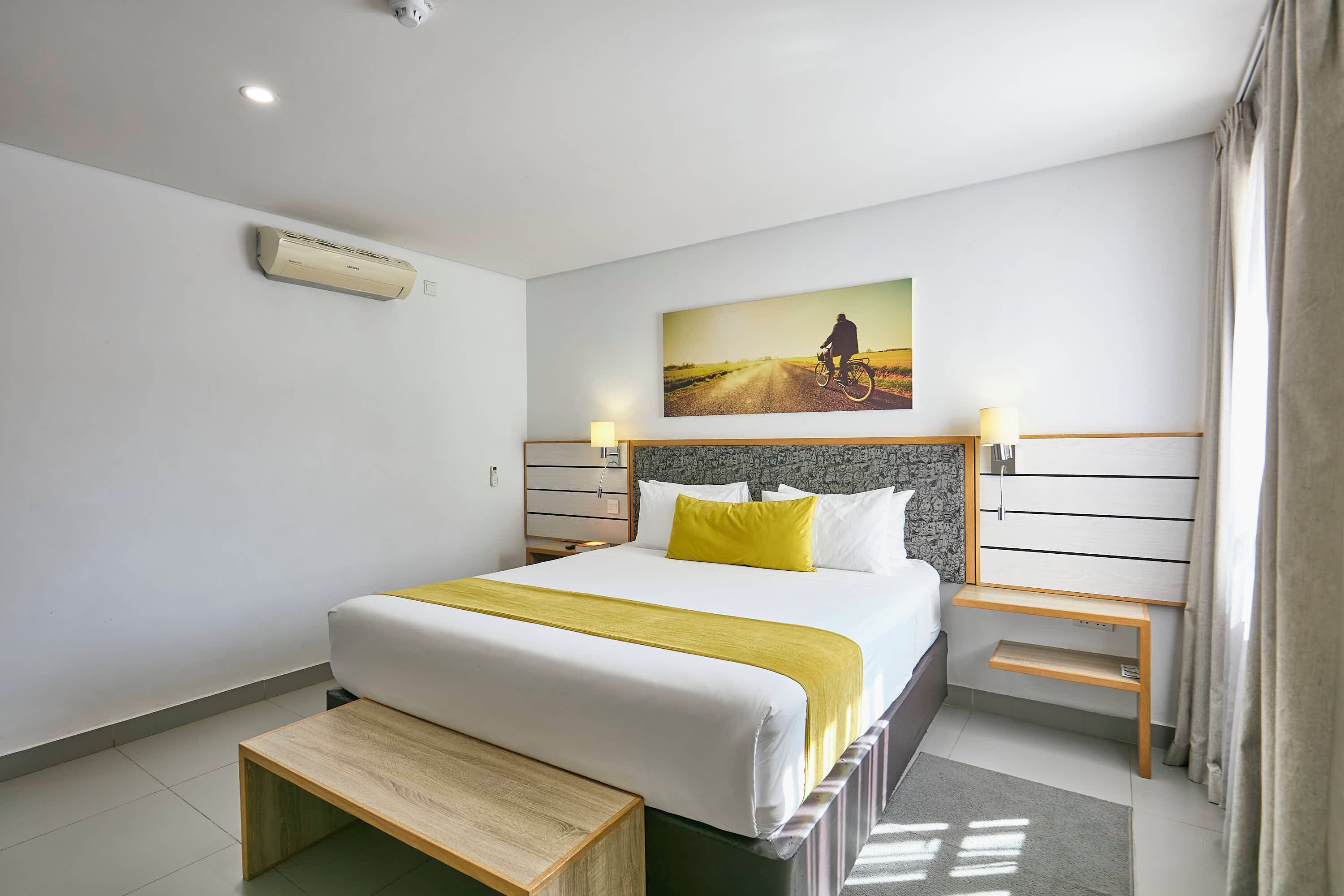 Quarto de hotel com uma cama grande, mesa de cabeceira, um banco, cabeceira, ar condicionado fixado na parede, um quadro e uma janela com cortinas.
