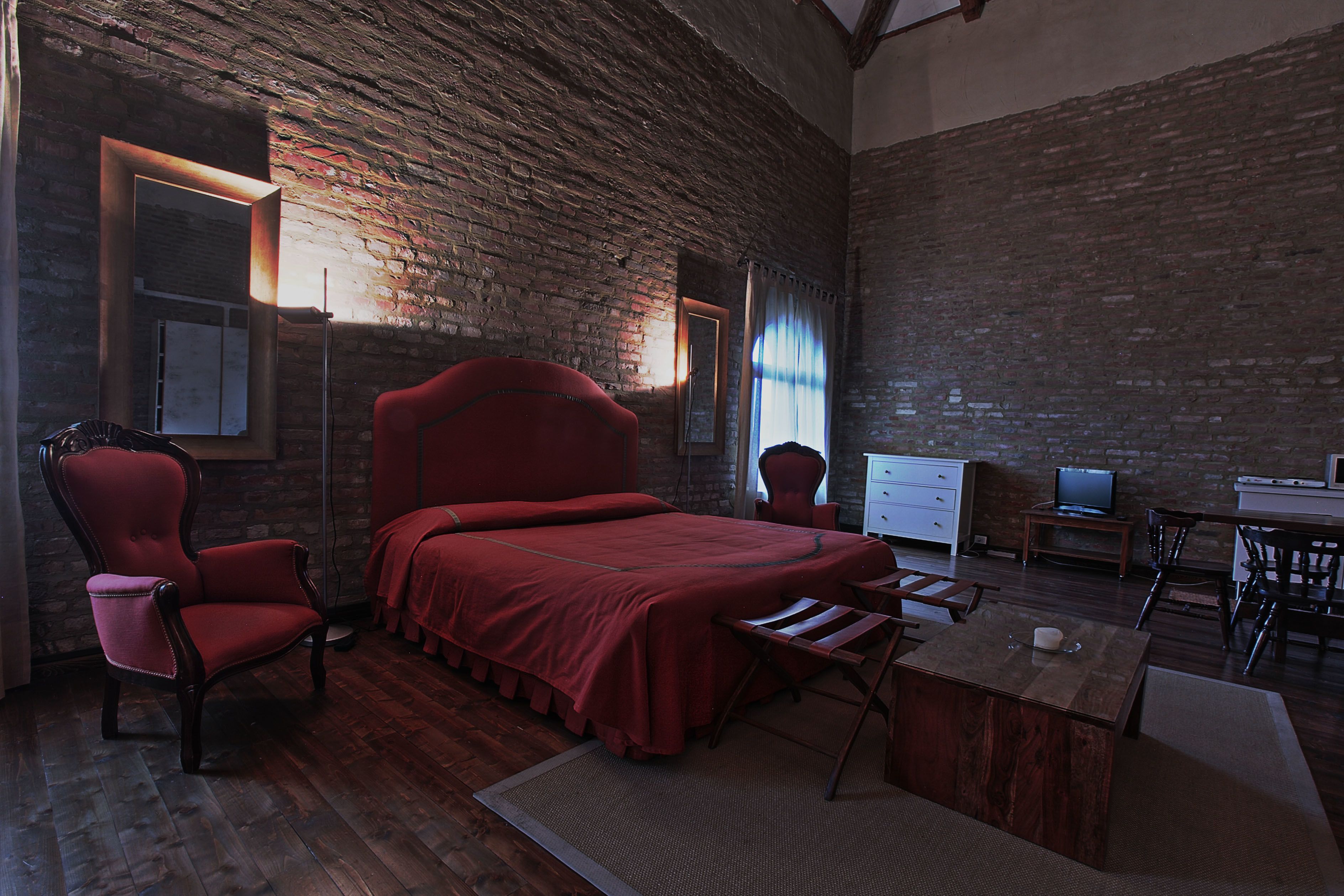 Quarto com parede de tijolos, cama grande, escrivaninha e uma cadeira.