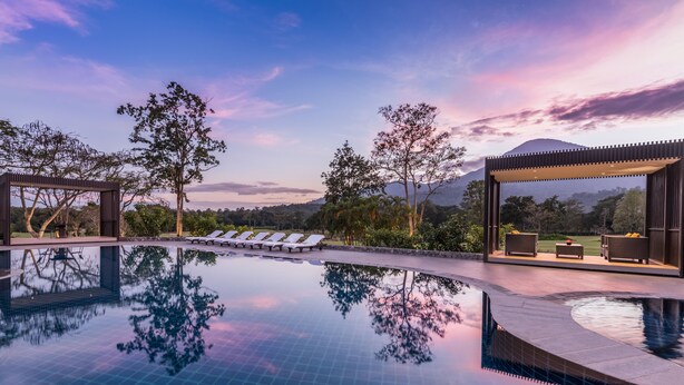 Uma piscina com espreguiçadeiras e área de estar coberta, cercada por árvores e um cenário montanhoso.