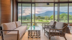 Quarto moderno com sofá, poltrona e mesa, grandes janelas com vista para um campo de golfe e montanhas.