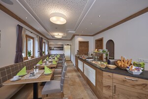 Uma longa área de refeições com bancos e mesas de madeira, um balcão de buffet com pão e doces, e vista para o exterior por grandes janelas.