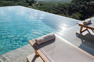 Uma piscina infinita com águas cristalinas azuis, rodeada por vegetação exuberante e um deck de madeira com espreguiçadeiras.