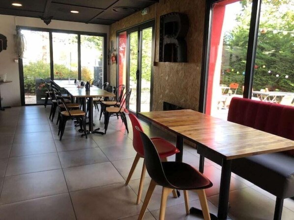 Interior de restaurante com uma longa mesa de madeira e cadeiras, janelas amplas e um assento em vermelho.