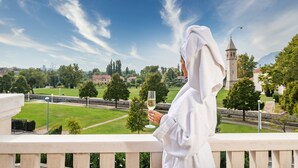 Uma pessoa vestida com bata branca segura um copo de vinho branco, admirando um parque com uma torre ao fundo.