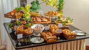 Um buffet com diversos produtos de panificação e pastelaria, incluindo bolos, biscoitos e pão, disposto numa bancada com uma planta ao fundo.