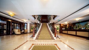 Um amplo hall de hotel com escadaria central, balcão de recepção e áreas de estar.