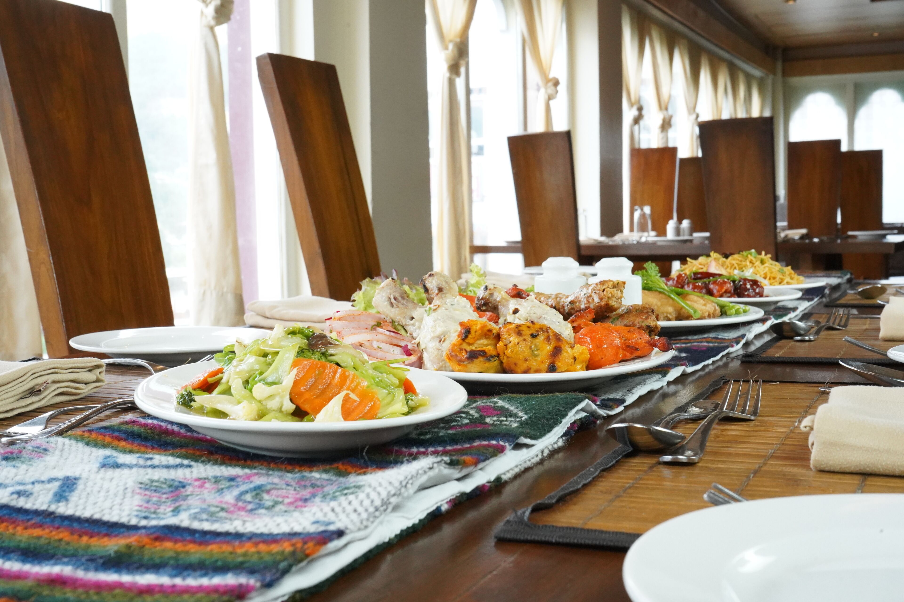 Mesa de jantar posta com diversos pratos, incluindo saladas e carnes, num restaurante com cadeiras de madeira e um jogo americano estampado.