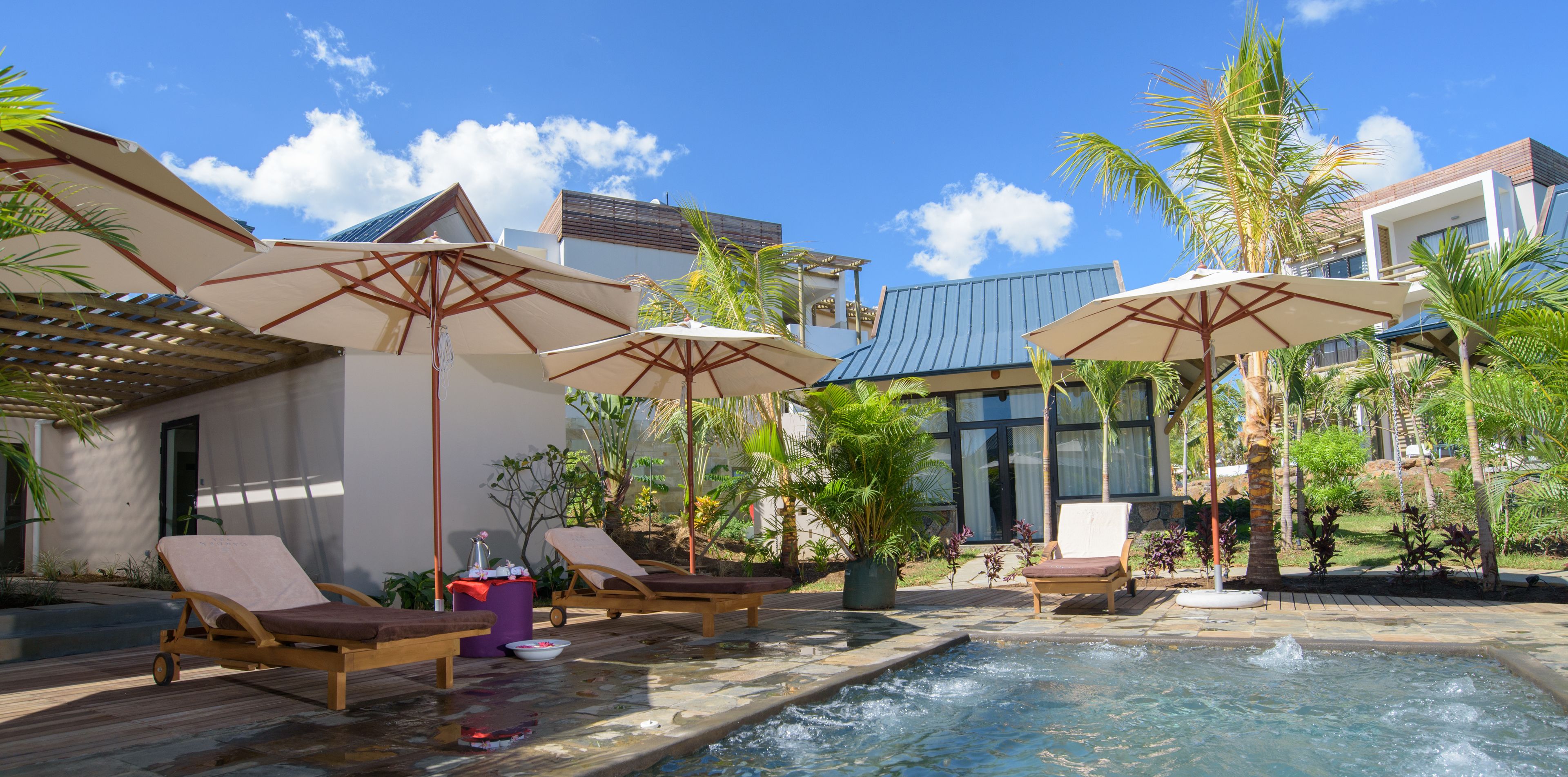 Uma área de piscina com espreguiçadeiras e guarda-sóis, cercada por plantas tropicais e arquitetura moderna.