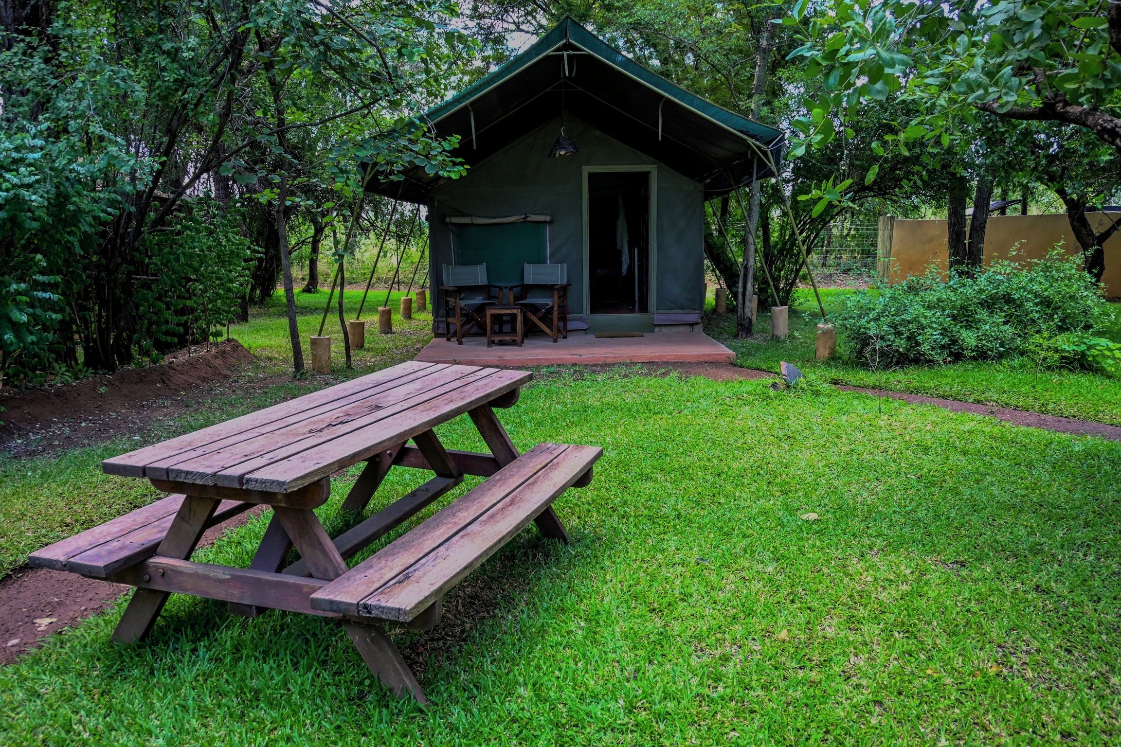 Uma mesa de piquenique de madeira com bancos em uma área gramada, com um pequeno edifício ao fundo.
