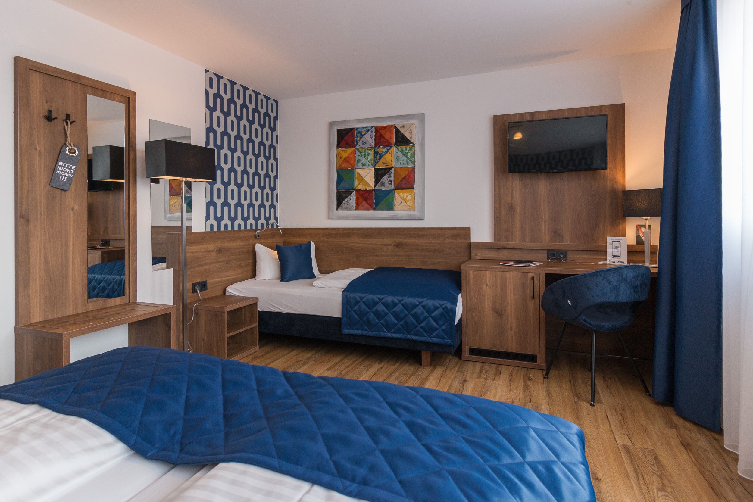 Quarto de hotel com cama, escrivaninha, cadeira, televisão, espelho e uma obra de arte colorida na parede.