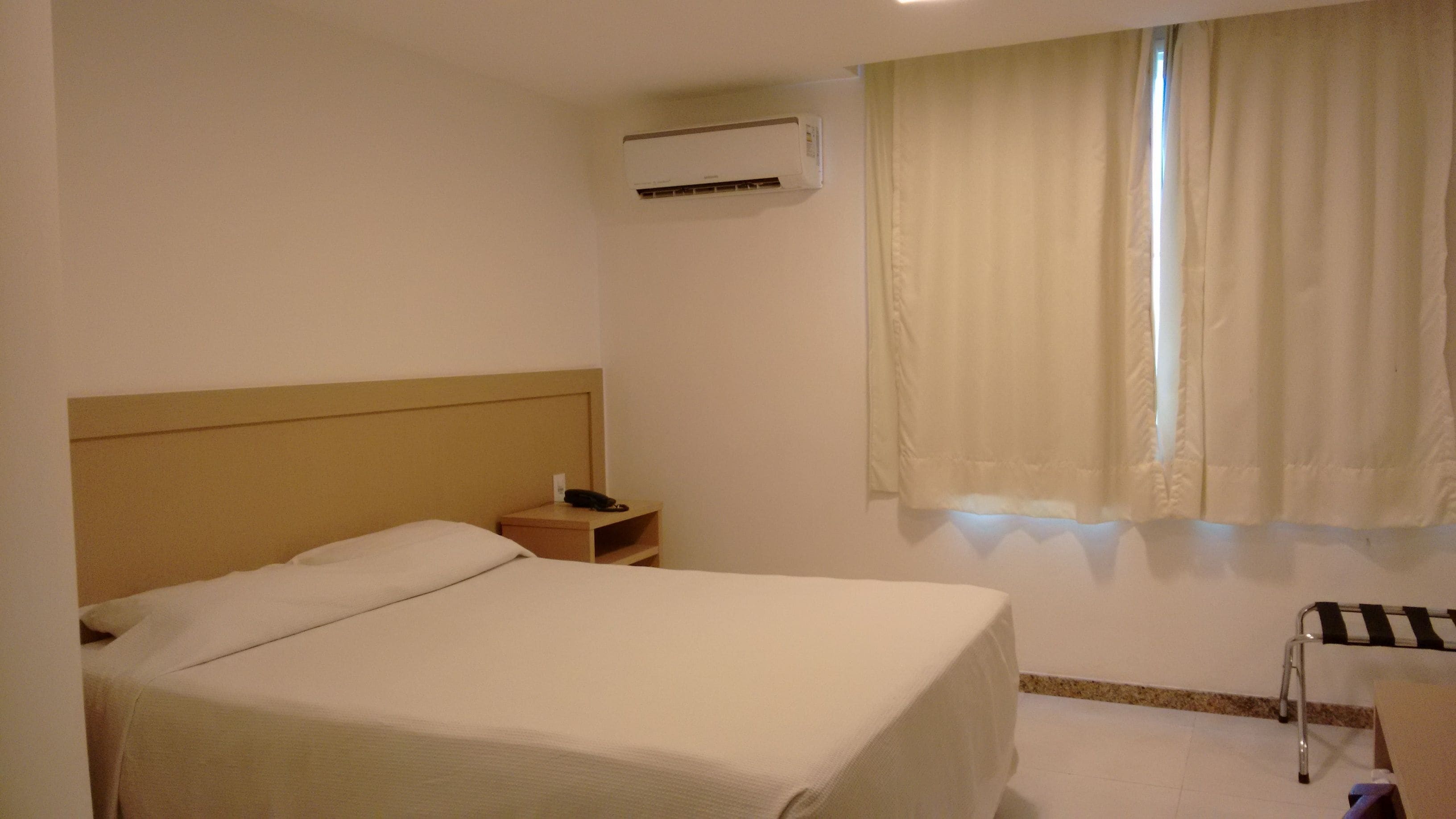 Quarto de hotel com cama, mesa de cabeceira, aparelho de ar condicionado, janela com cortinas e uma cadeira de rodas.