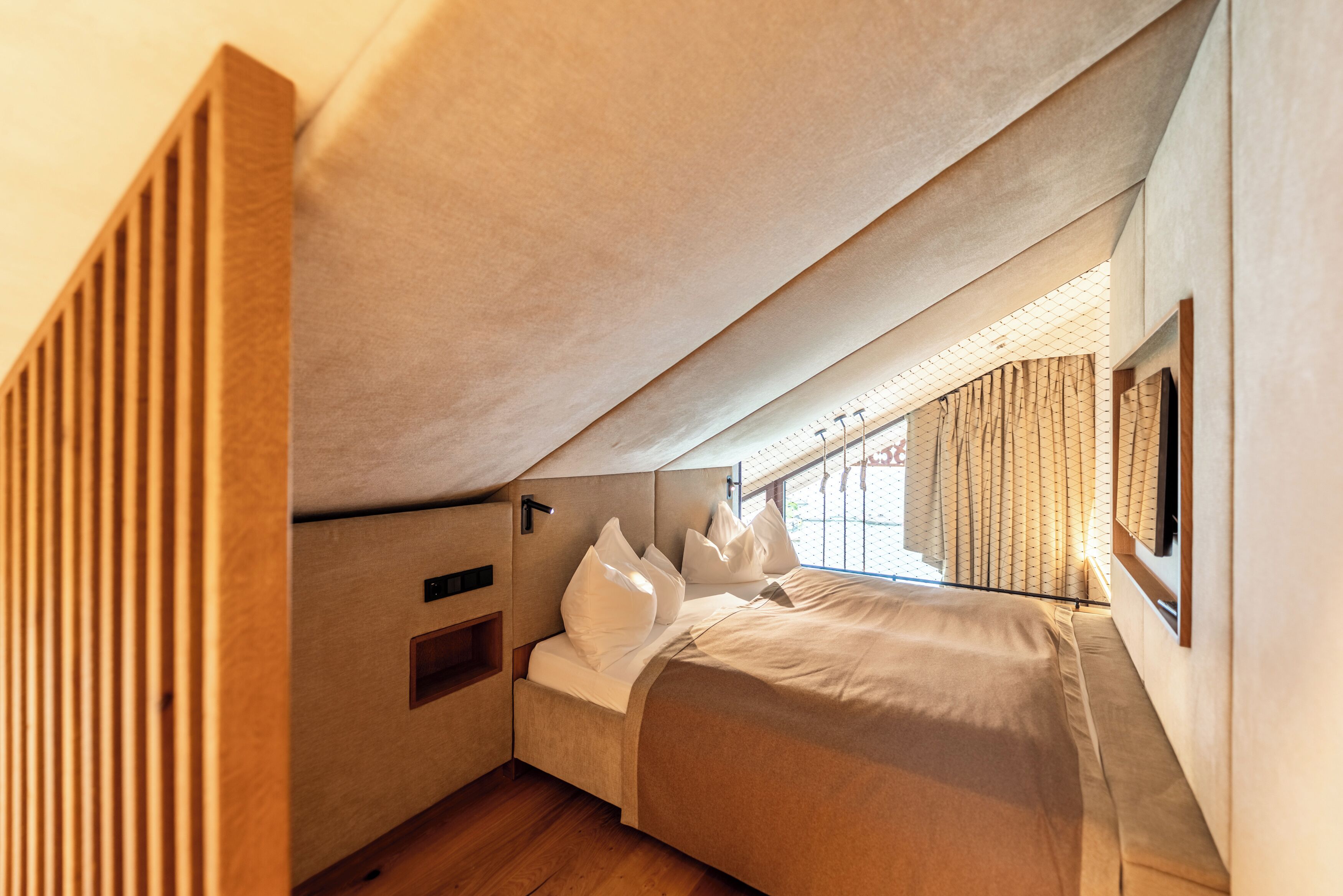 Quarto com teto inclinado, cama com roupa de cama branca, cabeceira de madeira e janela com vista.