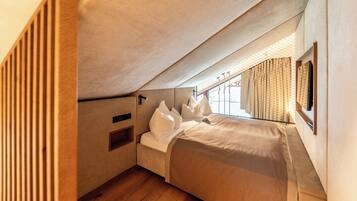Quarto com teto inclinado, cama com roupa de cama branca, cabeceira de madeira e janela com vista.