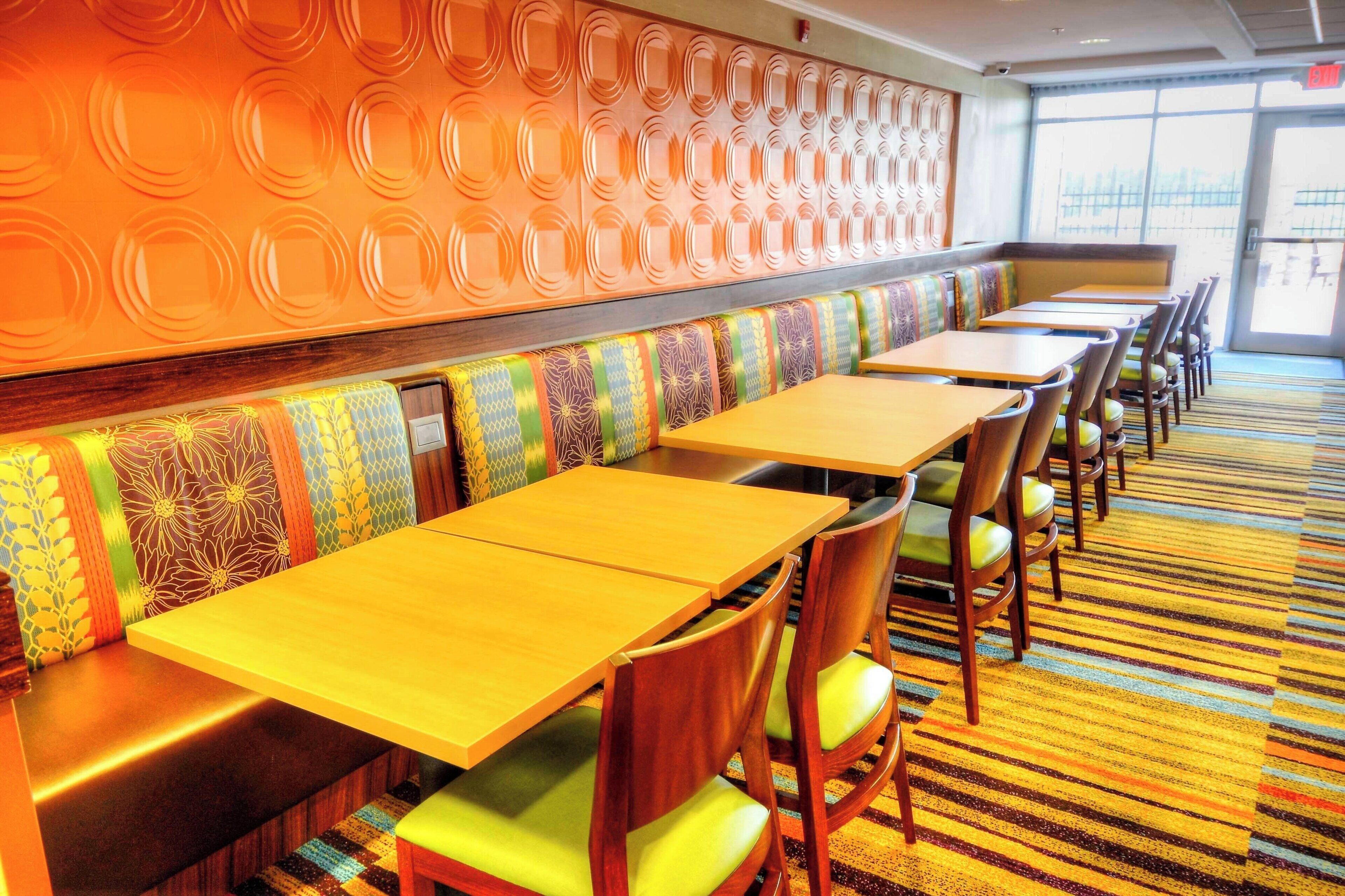 Um restaurante com mesas de madeira compridas e assentos acolchoados, parede estampada e carpete listrado e colorido.