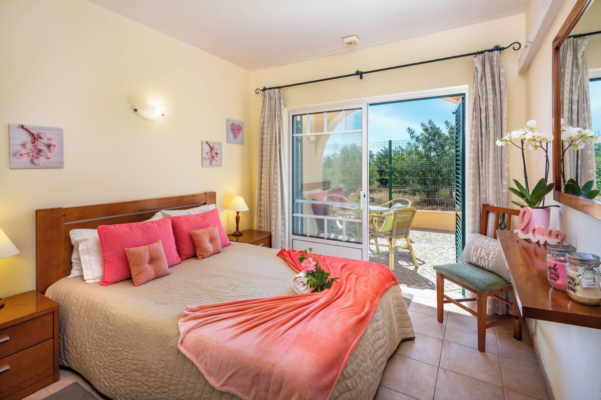 Quarto bem iluminado com uma cama grande, uma mesinha de cabeceira de madeira, um criado-mudo com abajur e vista para o exterior por uma porta de vidro deslizante.