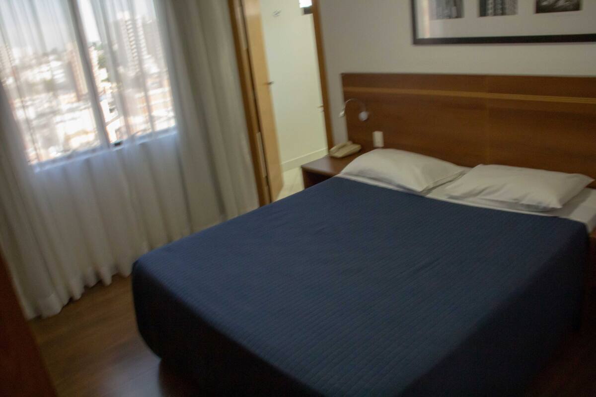 Quarto de hotel com cama, mesa de cabeceira, telefone e janela com cortinas.