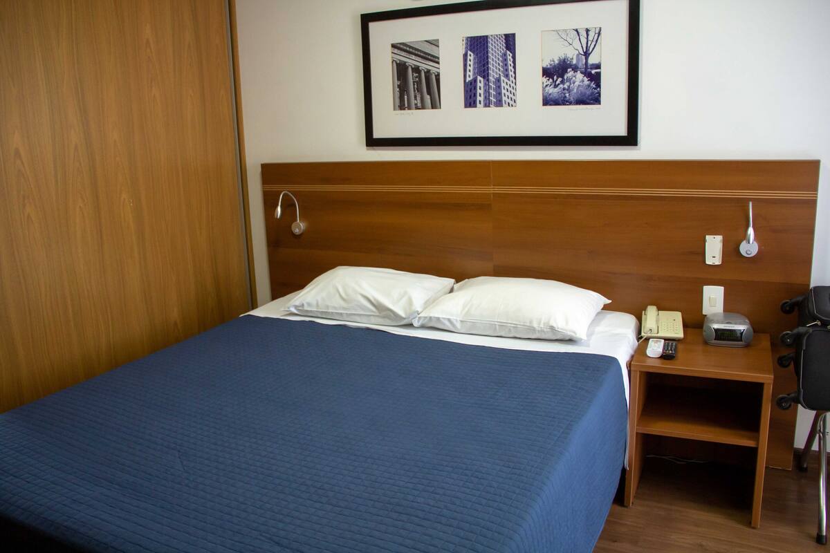 Quarto de hotel com cabeceira de madeira, cama com colcha azul, duas almofadas brancas, mesa de cabeceira com telefone, relógio pequeno e quadro emoldurado acima da cabeceira.
