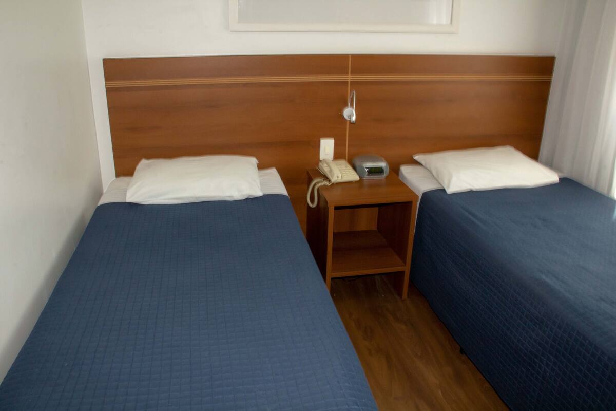 Quarto de hotel com duas camas de solteiro, cada uma com cabeceira de madeira, mesa de cabeceira com telefone e relógio digital, e uma parede branca lisa.