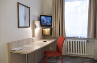 Quarto de hotel com uma escrivaninha, televisão, uma cadeira vermelha, um quadro na parede e uma janela com cortinas.