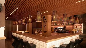 Uma área de bar com balcão de madeira, várias luminárias pendentes e uma variedade de garrafas e copos nas prateleiras.