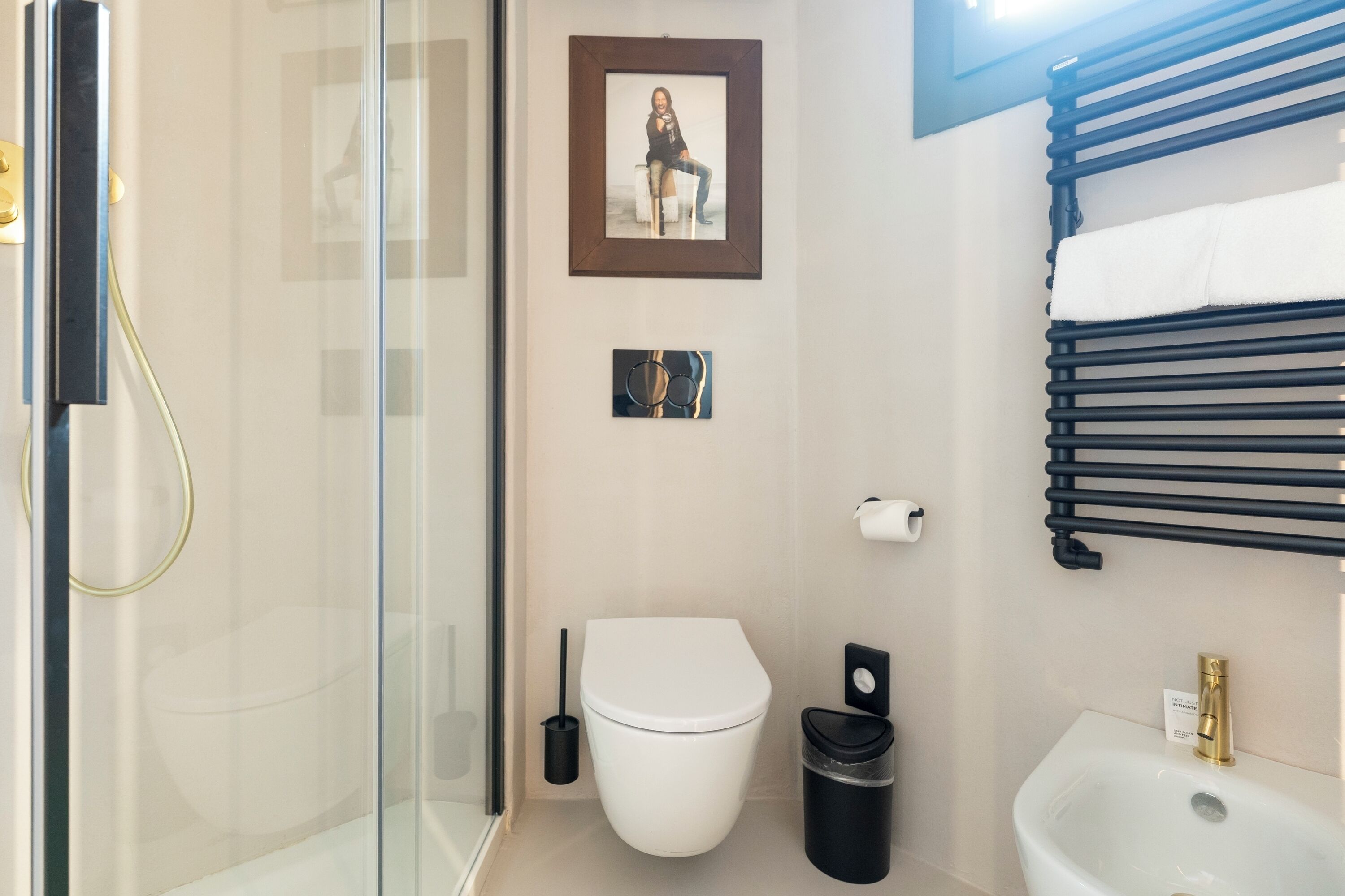 Uma casa de banho moderna com divisória de chuveiro em vidro, sanita branca, lavatório com torneira dourada, um cesto de lixo preto e uma fotografia emoldurada na parede.