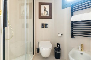 Uma casa de banho moderna com divisória de chuveiro em vidro, sanita branca, lavatório com torneira dourada, um cesto de lixo preto e uma fotografia emoldurada na parede.