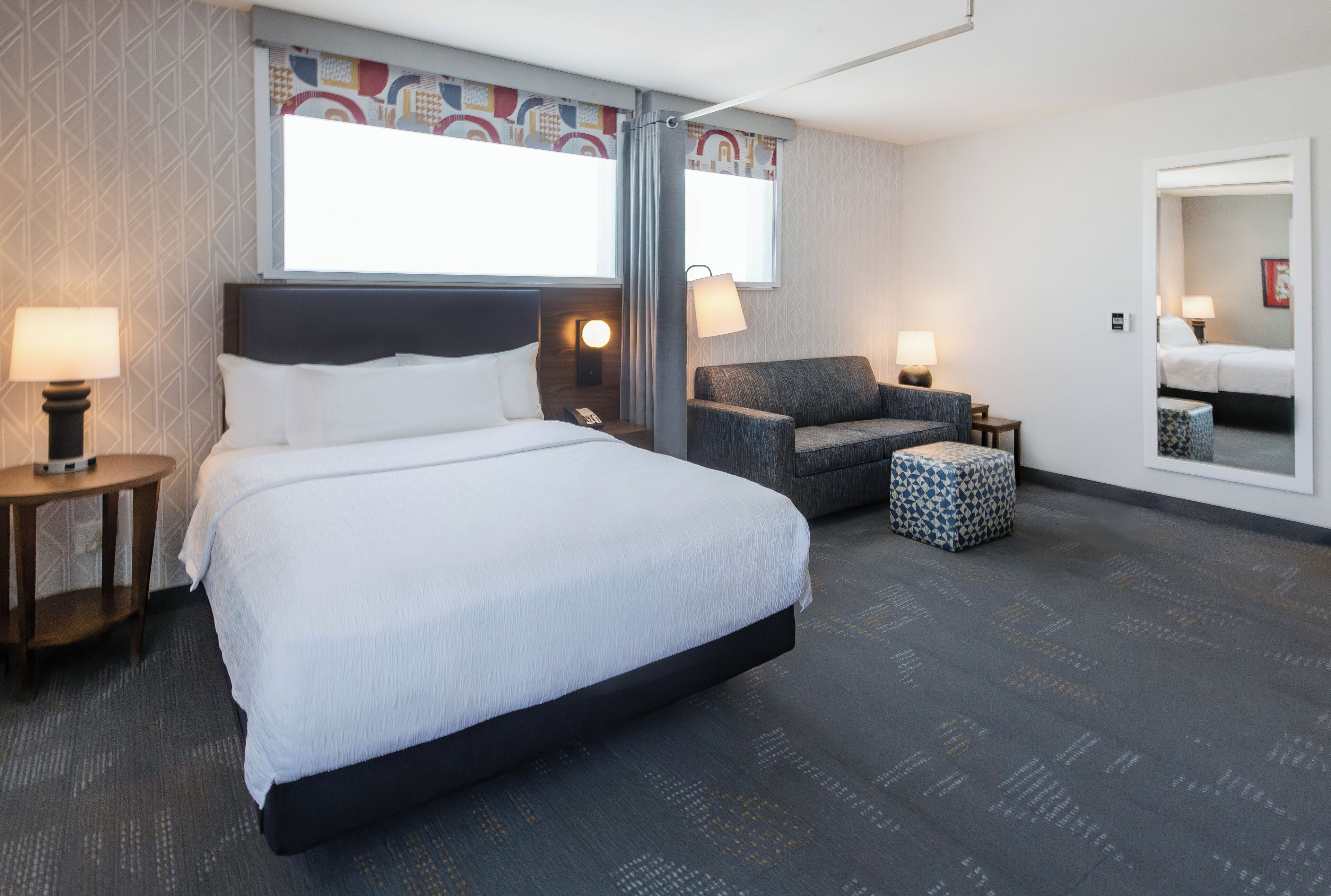 Quarto de hotel com uma cama grande, uma cadeira, uma mesinha, uma luminária e um espelho.