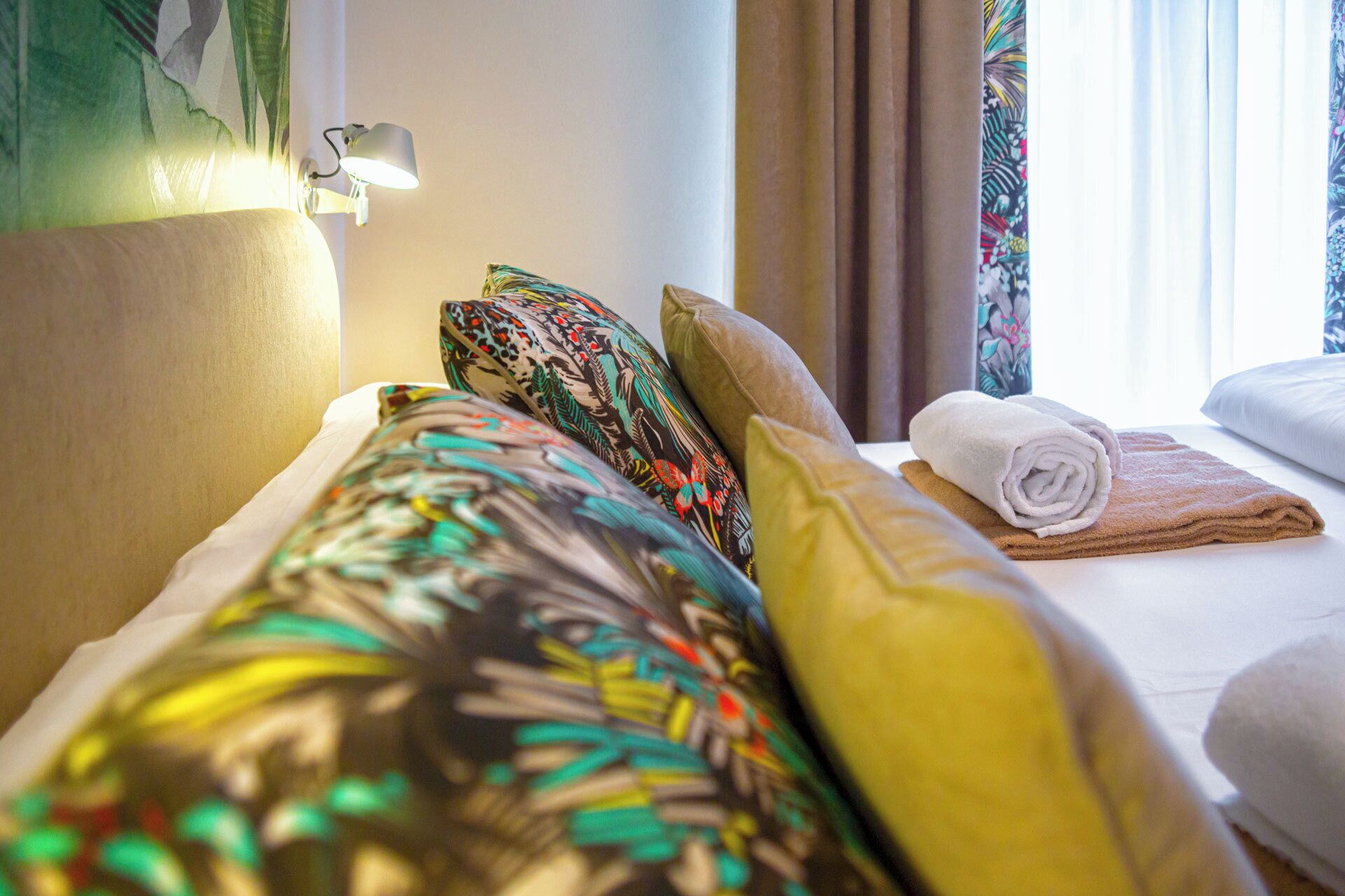 Uma cama bem arrumada com almofadas coloridas, uma toalha enrolada e um abajur na mesinha de cabeceira.