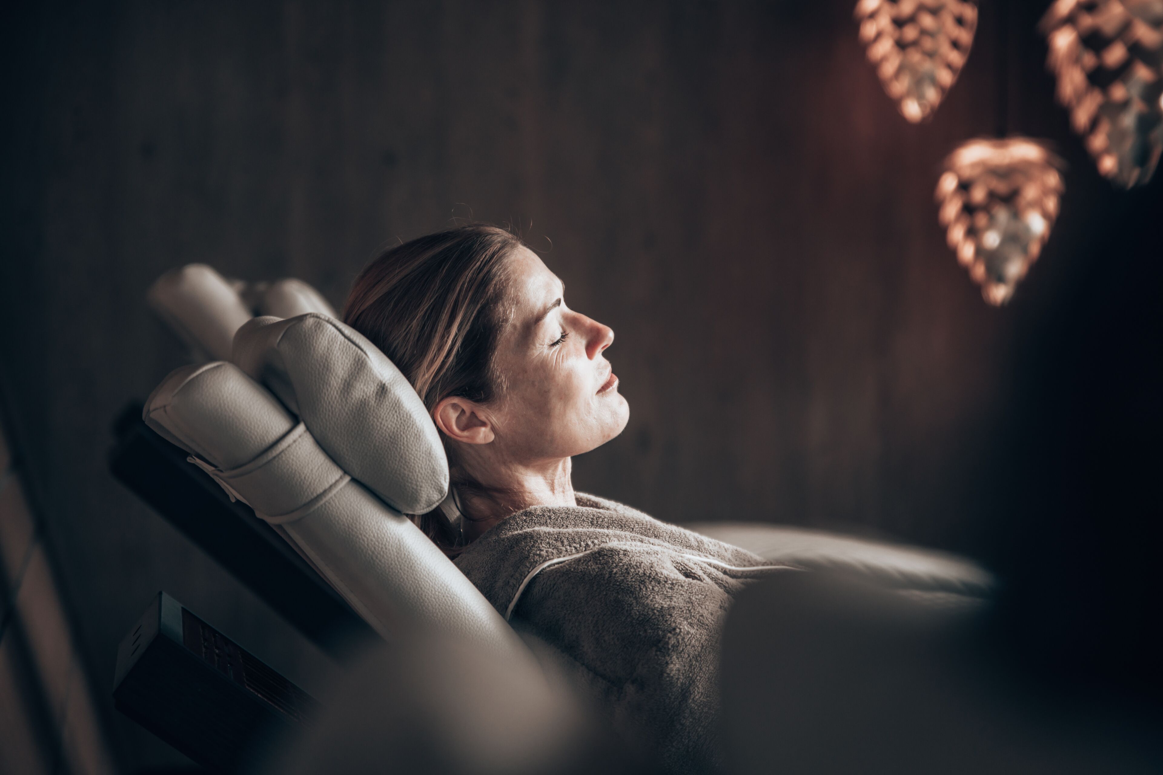 Uma pessoa relaxada em uma cadeira de massagem, com uma parede de madeira e luminárias pendentes em formato de coração ao fundo.