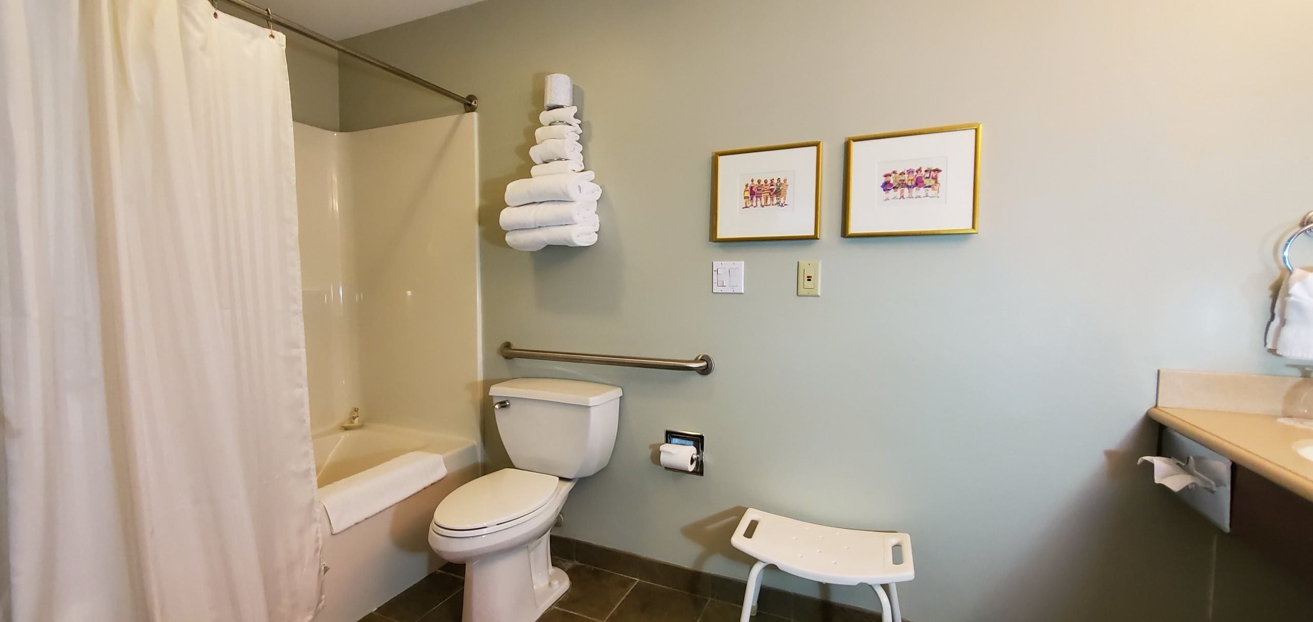 Uma casa de banho com chuveiro, sanita e um banquinho branco pequeno. Existem duas molduras com quadros na parede e um porta-toalhas com toalhas.