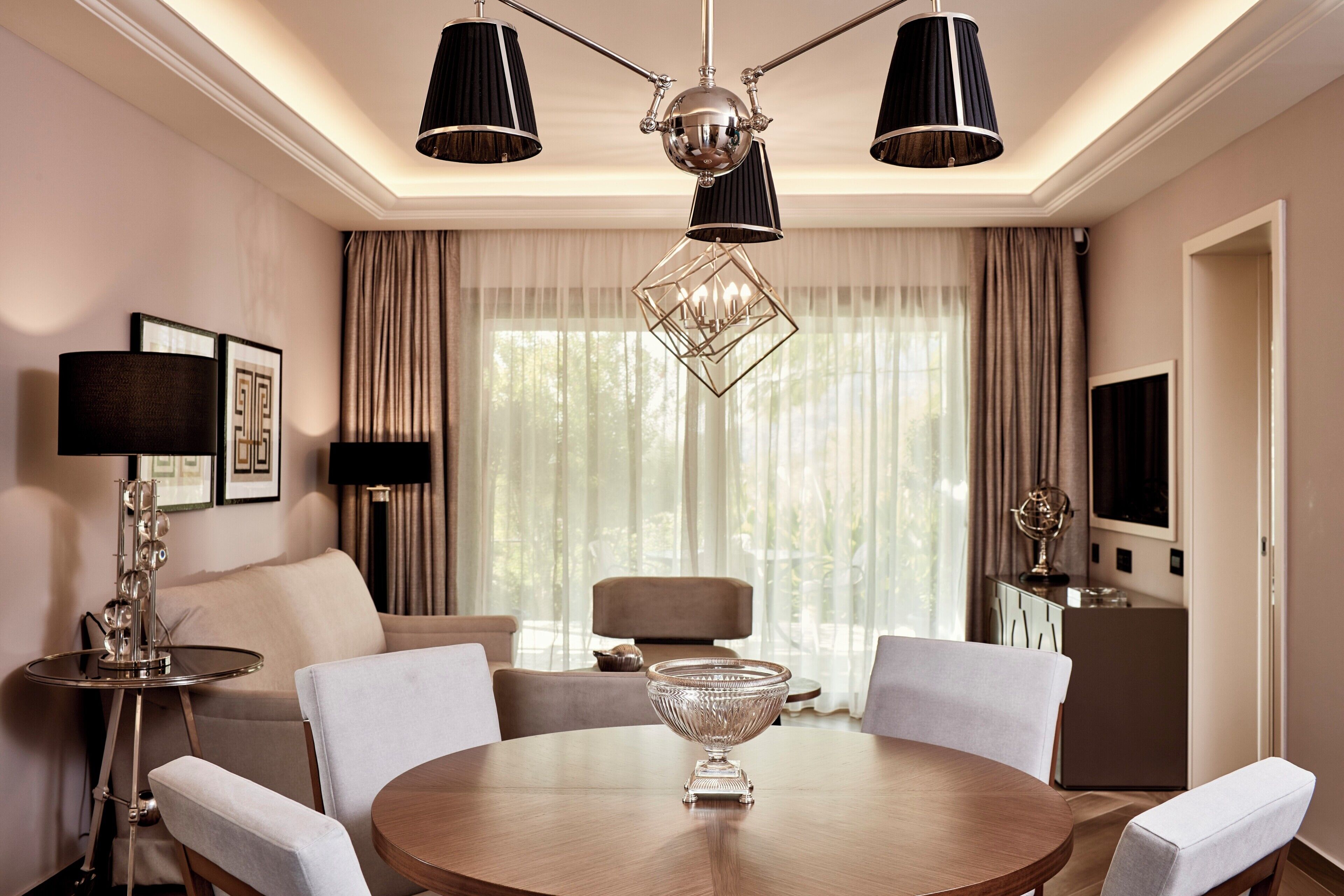 Quarto de hotel moderno com uma mesa de jantar redonda, um sofá, uma TV e um candelabro.