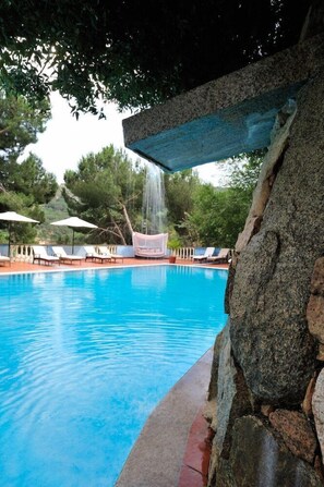 Piscina azul límpida com espreguiçadeiras e guarda-sóis, cercada por árvores e um paredão de rochas.