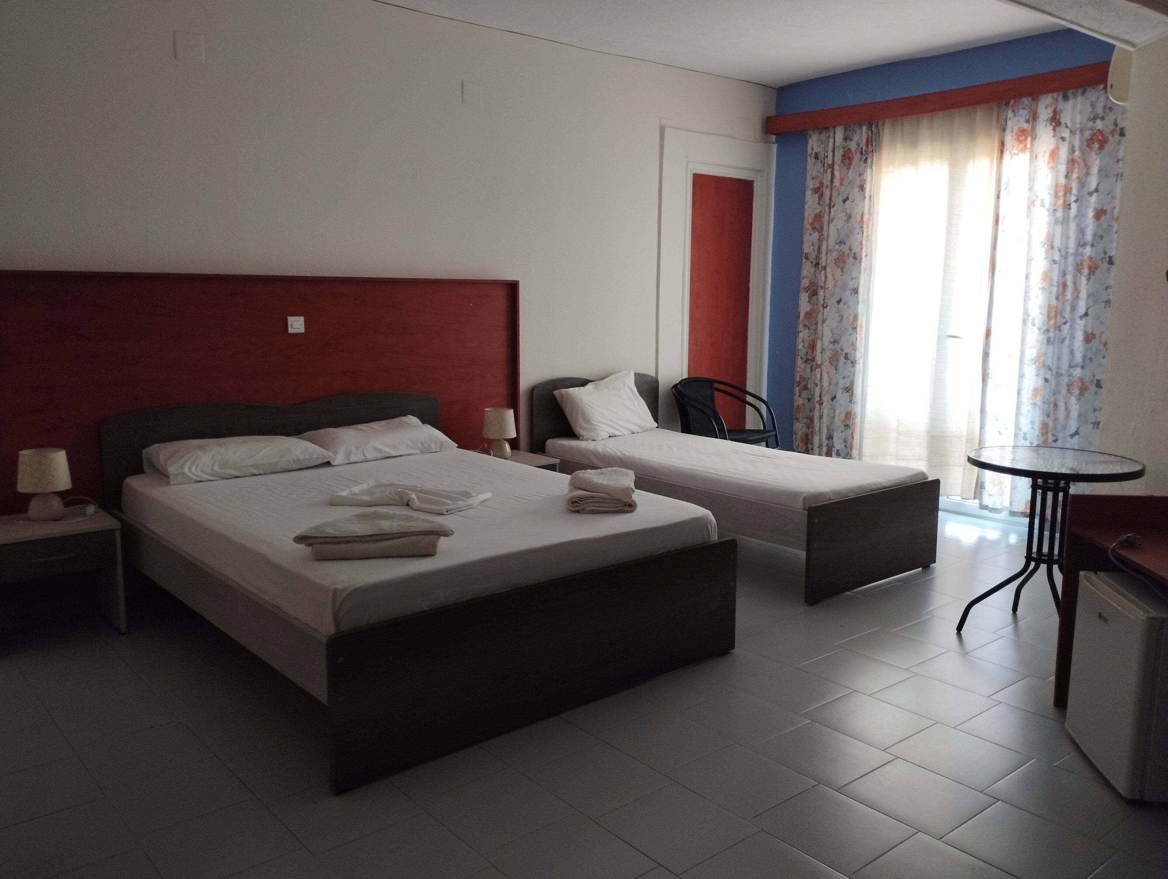 Quarto de hotel com duas camas, uma mesinha, uma cadeira e mesa de cabeceira com abajur.