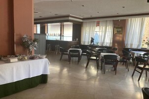 Uma área de refeições com mesas e cadeiras, um espaço de bar e vista para o exterior por grandes janelas.