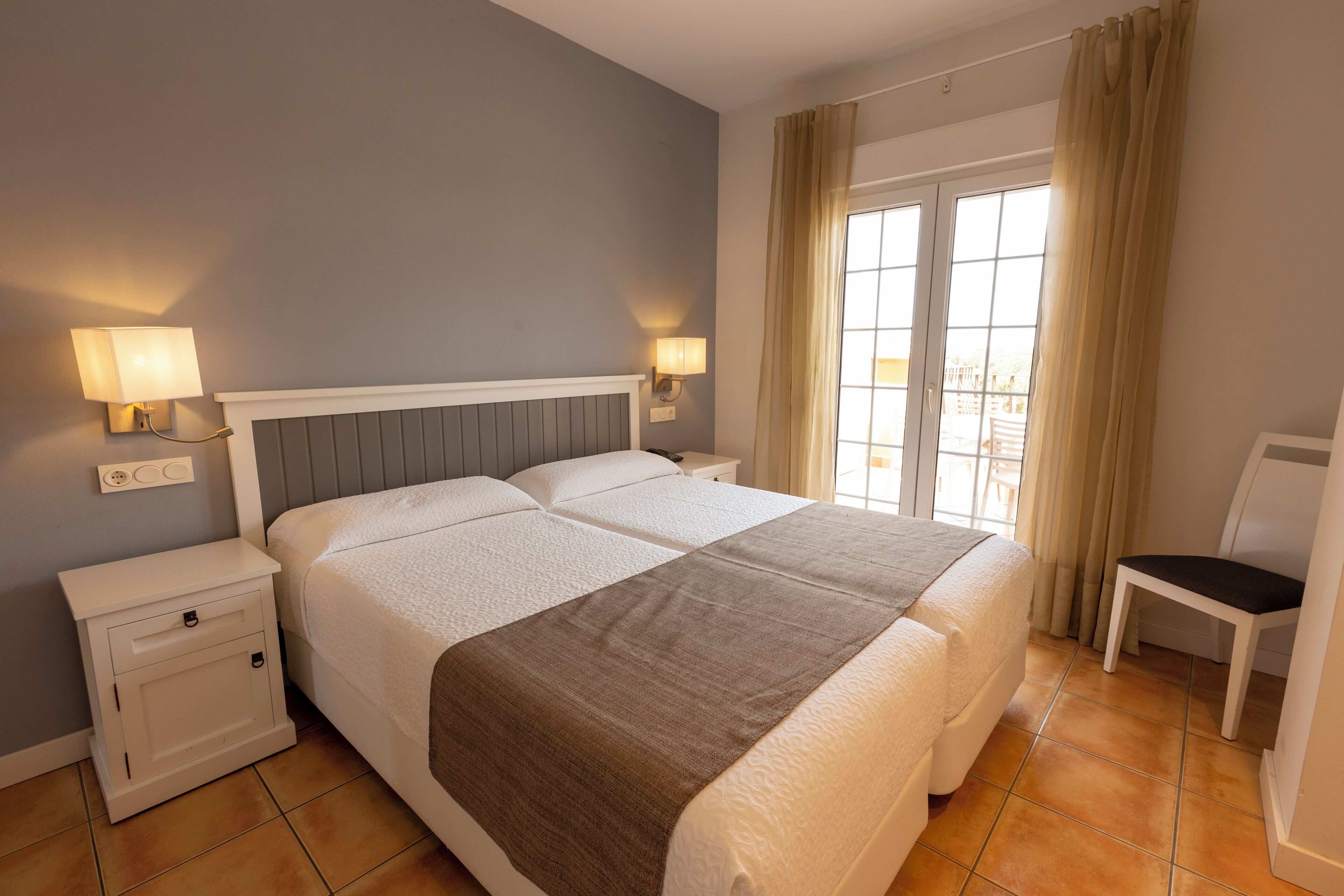 Quarto de hotel com uma cama grande, duas mesas de cabeceira, uma cadeira e uma janela com cortinas.