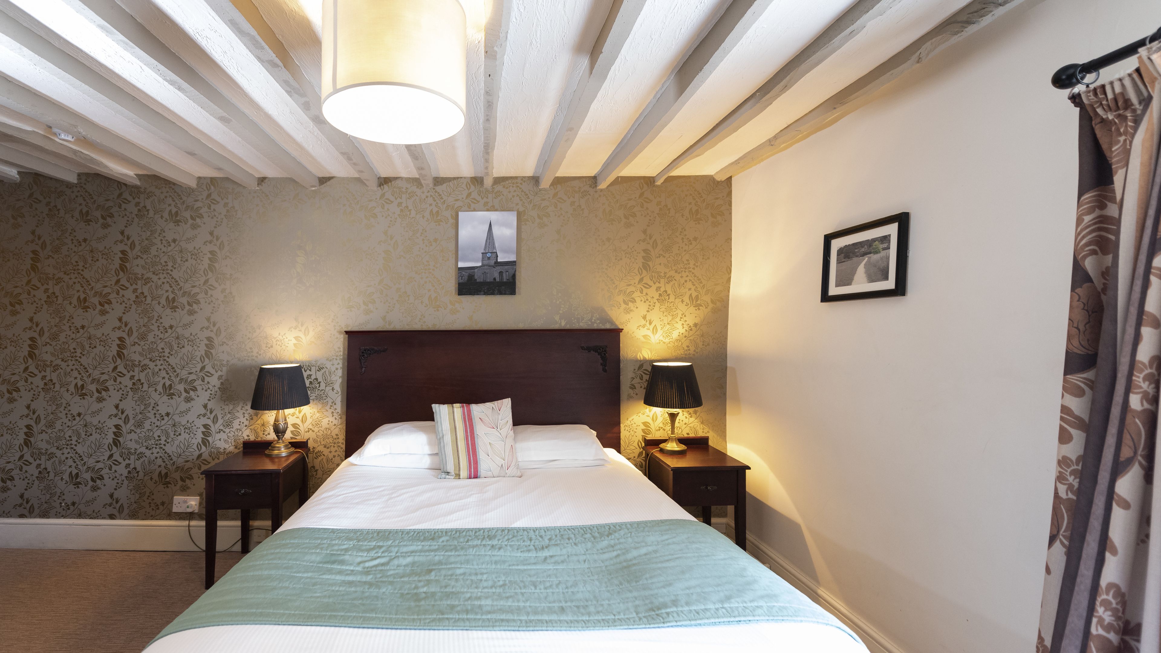 Quarto com uma cama grande, duas mesas de cabeceira com luminárias, papel de parede floral, teto de madeira e um quadro na parede.