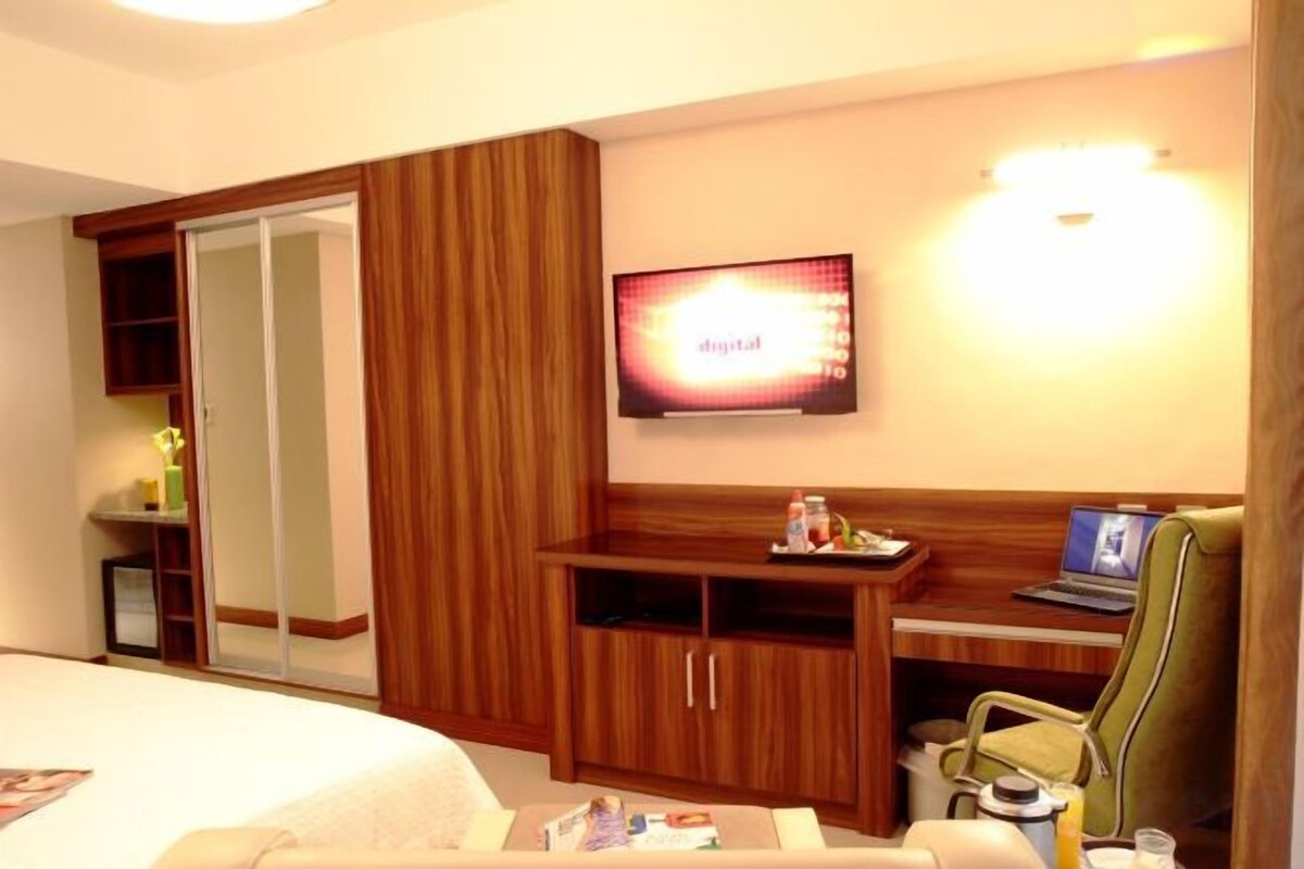 Quarto de hotel com cama, televisão na parede, secretária com um laptop e uma cadeira verde.