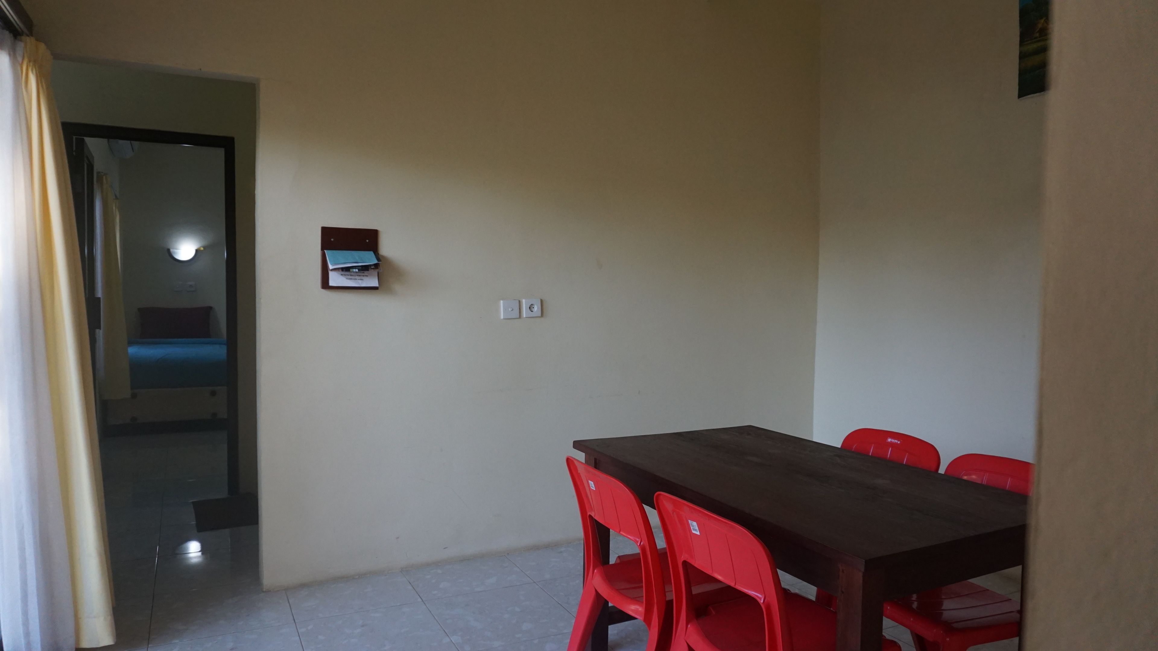Quarto com mesa de madeira e cadeiras vermelhas, porta que dá para outro ambiente e interruptor de luz fixado na parede.