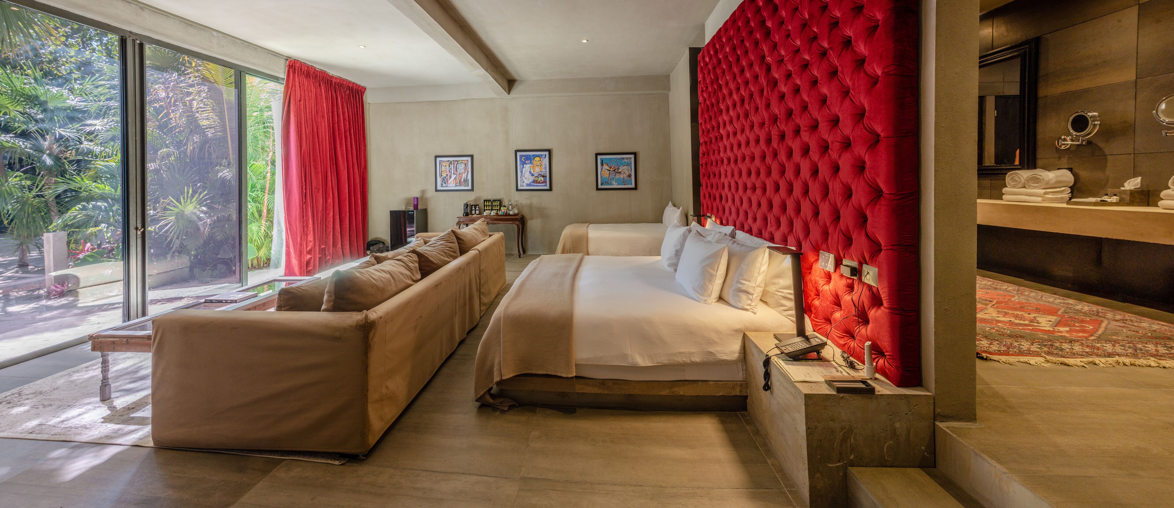 Quarto de hotel moderno com uma cama grande, um sofá, uma parede de destaque vermelha e vista para o verde lá fora.