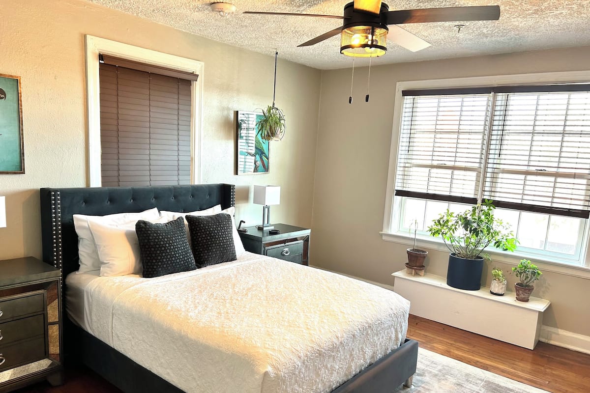 Quarto com cama grande, ventoinha no teto, janela com persianas e mesa de cabeceira com luminária.