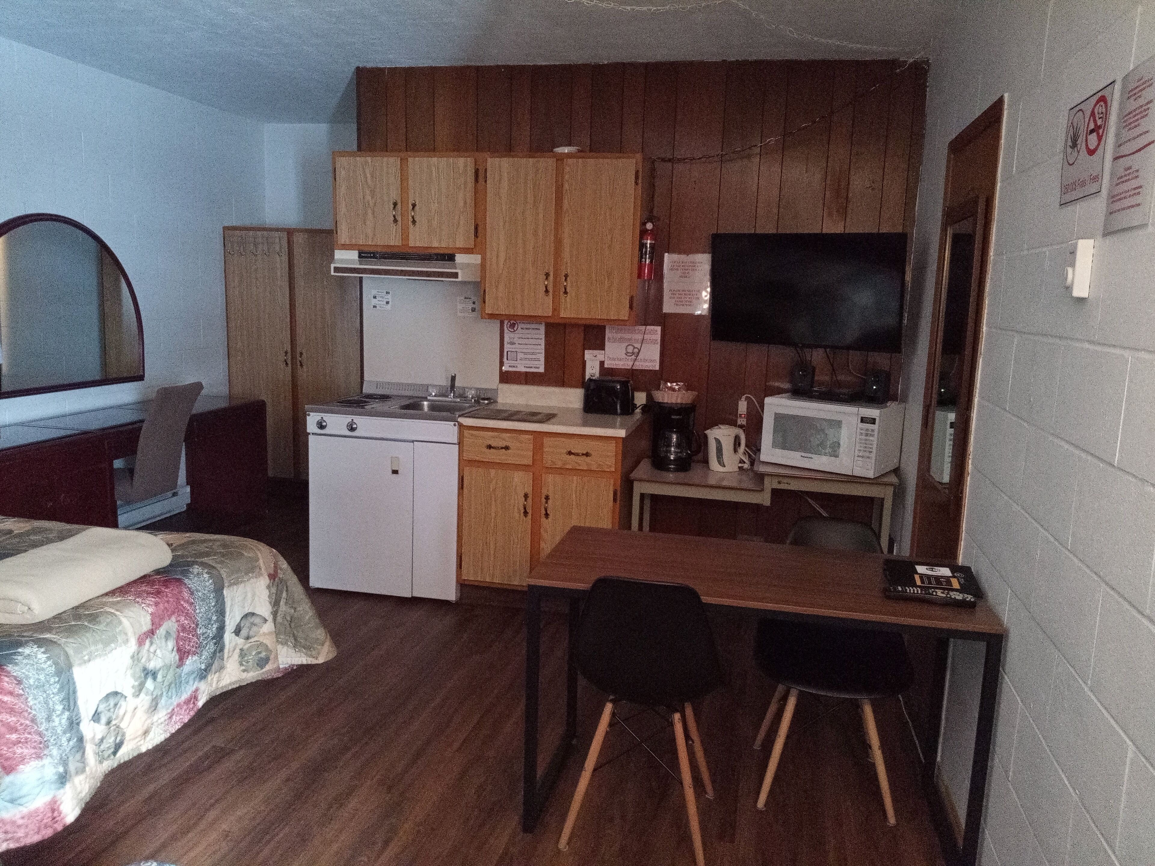 Quarto compacto com cozinha compacta, uma cama, escrivaninha com duas cadeiras e televisão.