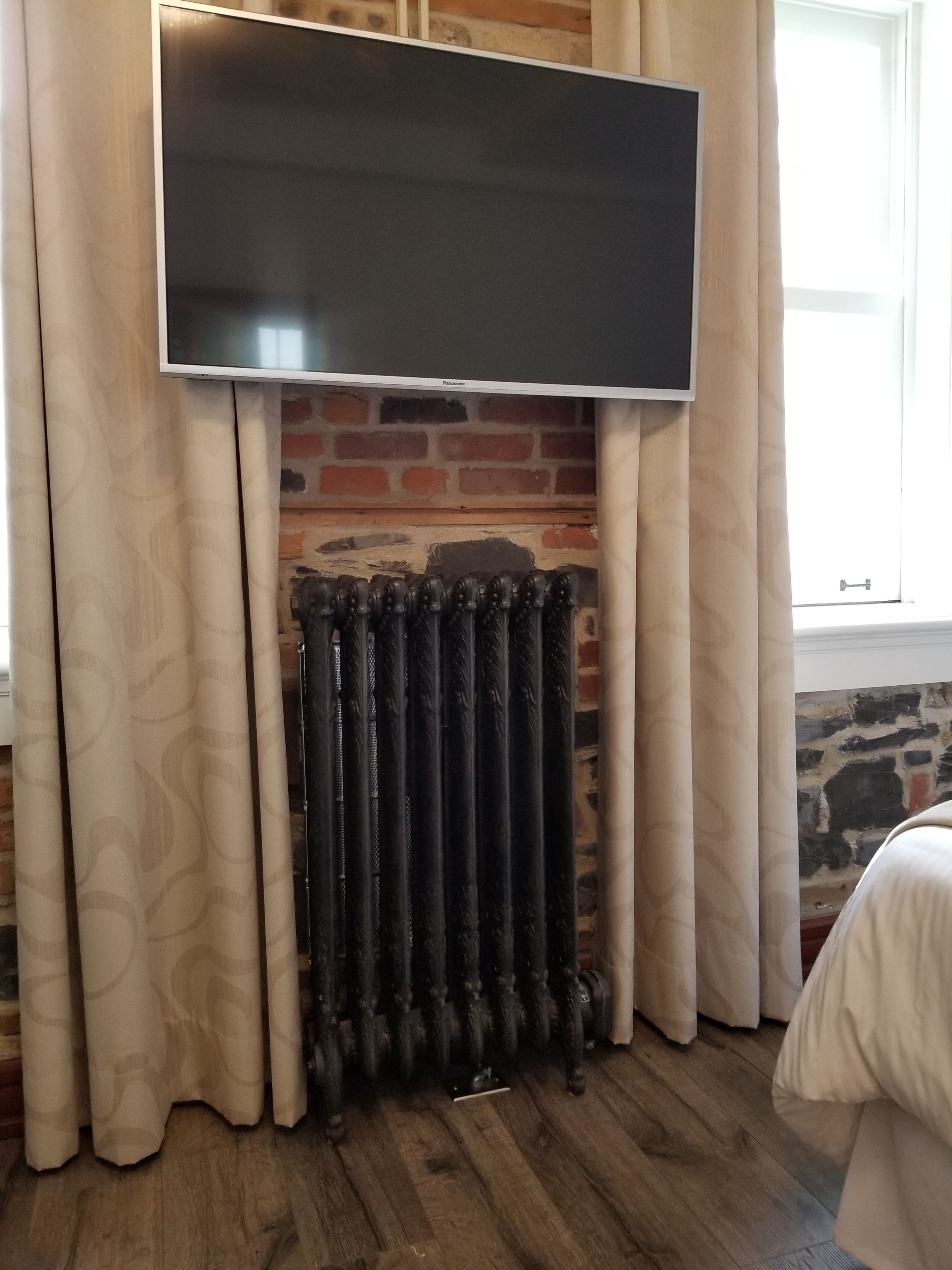 Quarto com televisão de tela plana presa a uma parede de tijolos, radiador abaixo e cortinas estampadas.