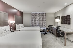 Quarto de hotel com duas camas, uma escrivaninha, uma cadeira, um abajur e cortina estampada.
