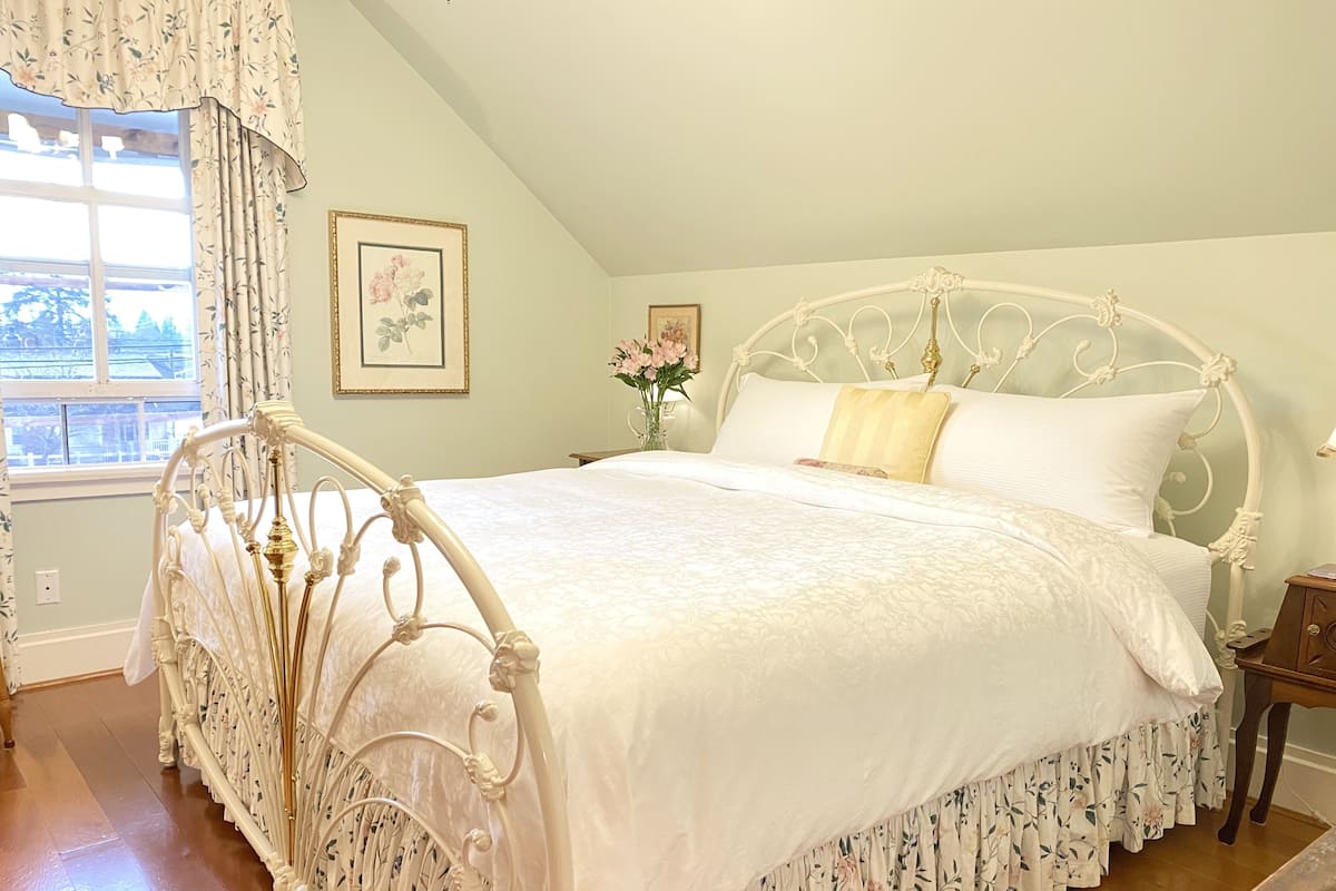 Quarto com cama branca ornamentada, cortinas florais, um quadro e vista pela janela.