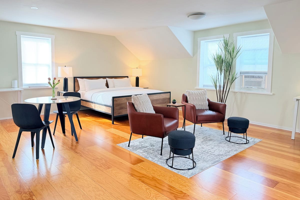 Um quarto moderno com uma cama, uma mesa de jantar, duas cadeiras, uma mesinha de apoio, um vaso com planta e uma janela grande.