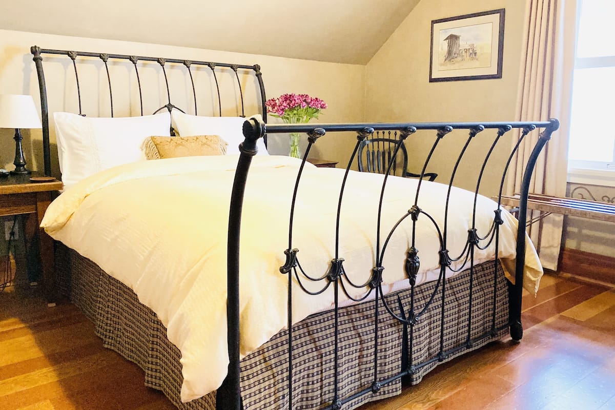 Uma cama bem arrumada com estrutura de metal, uma mesa de cabeceira com abajur e um quadro na parede.