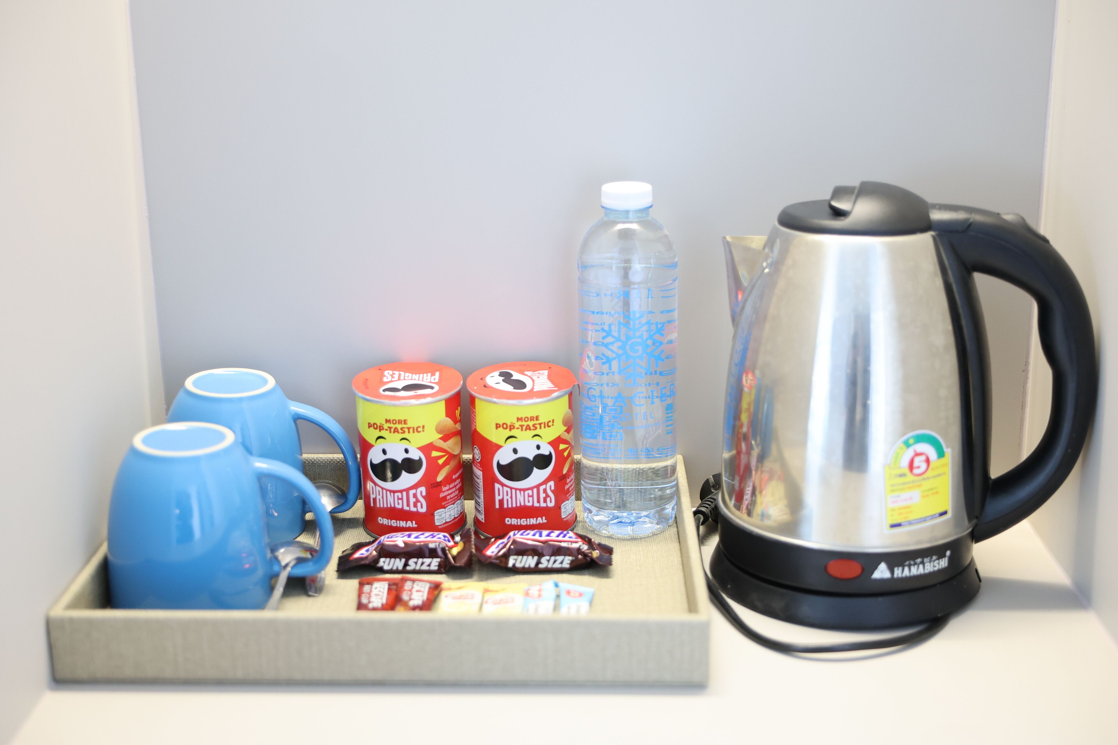 Uma prateleira com uma chaleira, duas canecas azuis, duas latas de Pringles, uma garrafa de água e alguns petiscos.
