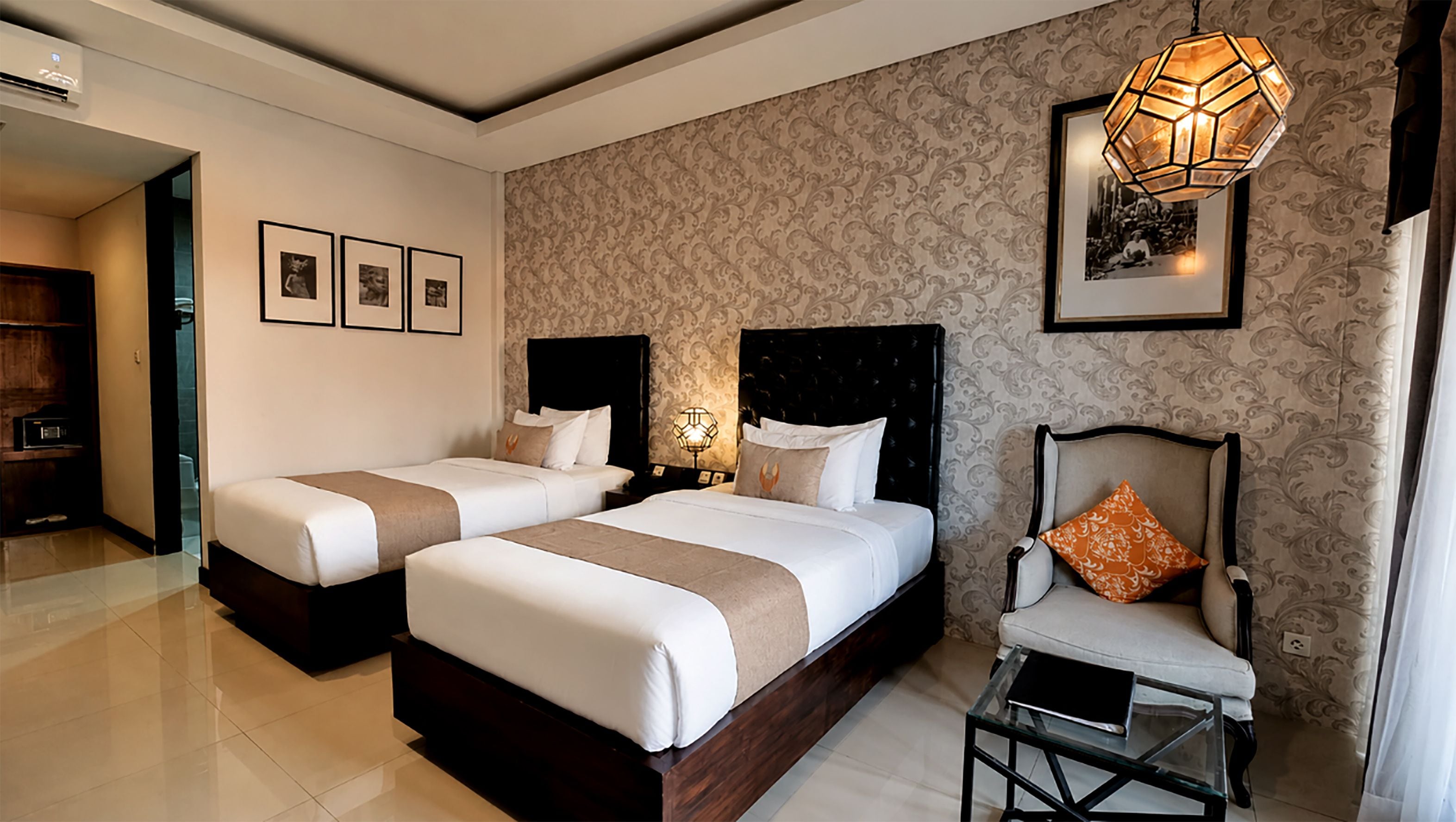 Quarto de hotel com duas camas, uma cadeira, mesa de cabeceira, abajur e quadros nas paredes.