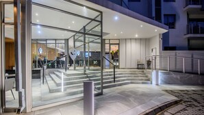 Entrada moderna de hotel com portas de vidro, escada e área externa bem iluminada.