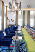 Um hall de hotel moderno com um sofá comprido, poltronas azuis e um sofá verde. Há mesas redondas e um tapete estampado.
